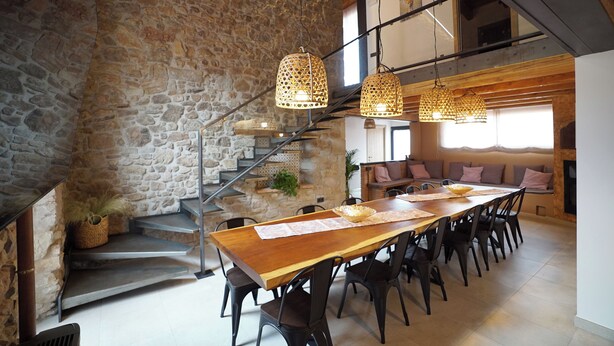 Uma sala de jantar com uma mesa de madeira comprida, cadeiras pretas e luminárias de vime suspensas. Uma parede de pedra e uma escadaria são visíveis ao fundo.
