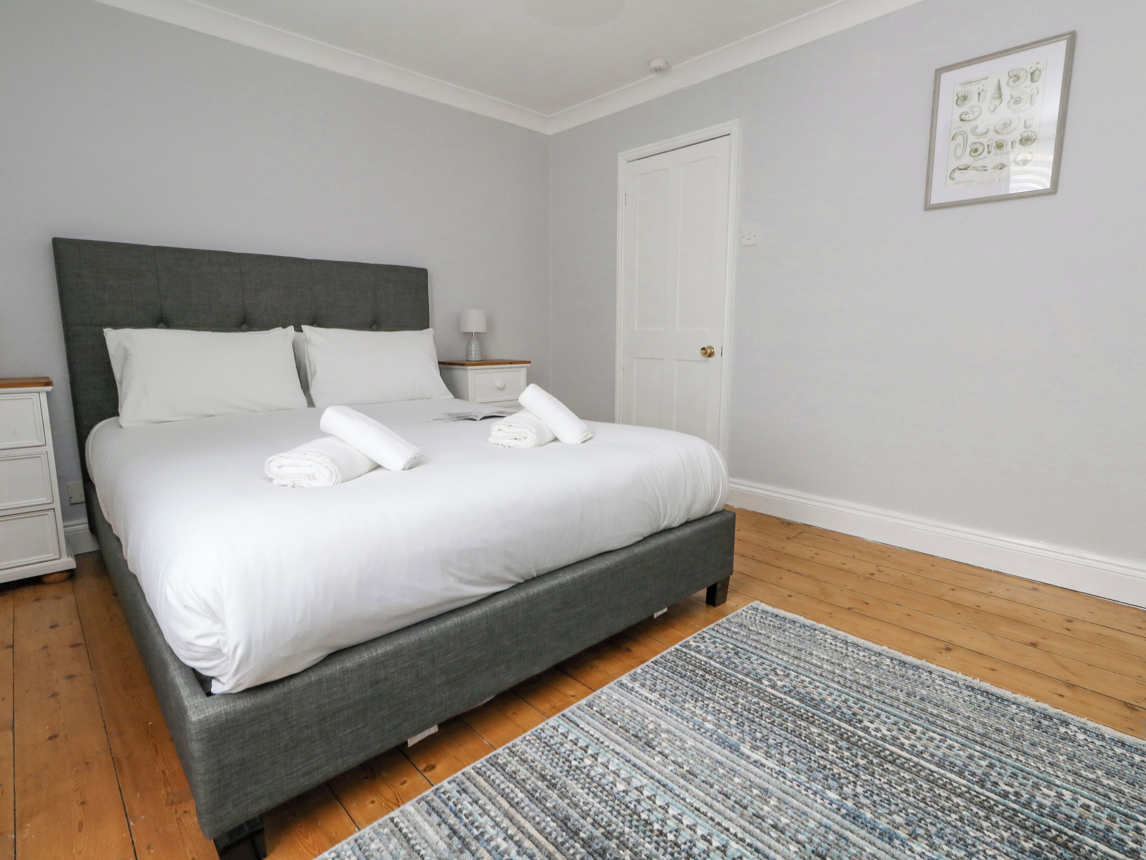 Uma cama bem arrumada com roupas de cama e toalhas brancas, uma cabeceira estofada cinza e piso de madeira.