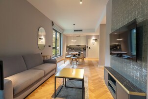 Uma sala de estar moderna com sofá, mesa de centro e televisão. Há uma área de jantar com cadeiras e mesa. O quarto possui janelas amplas e uma cozinha ao fundo.