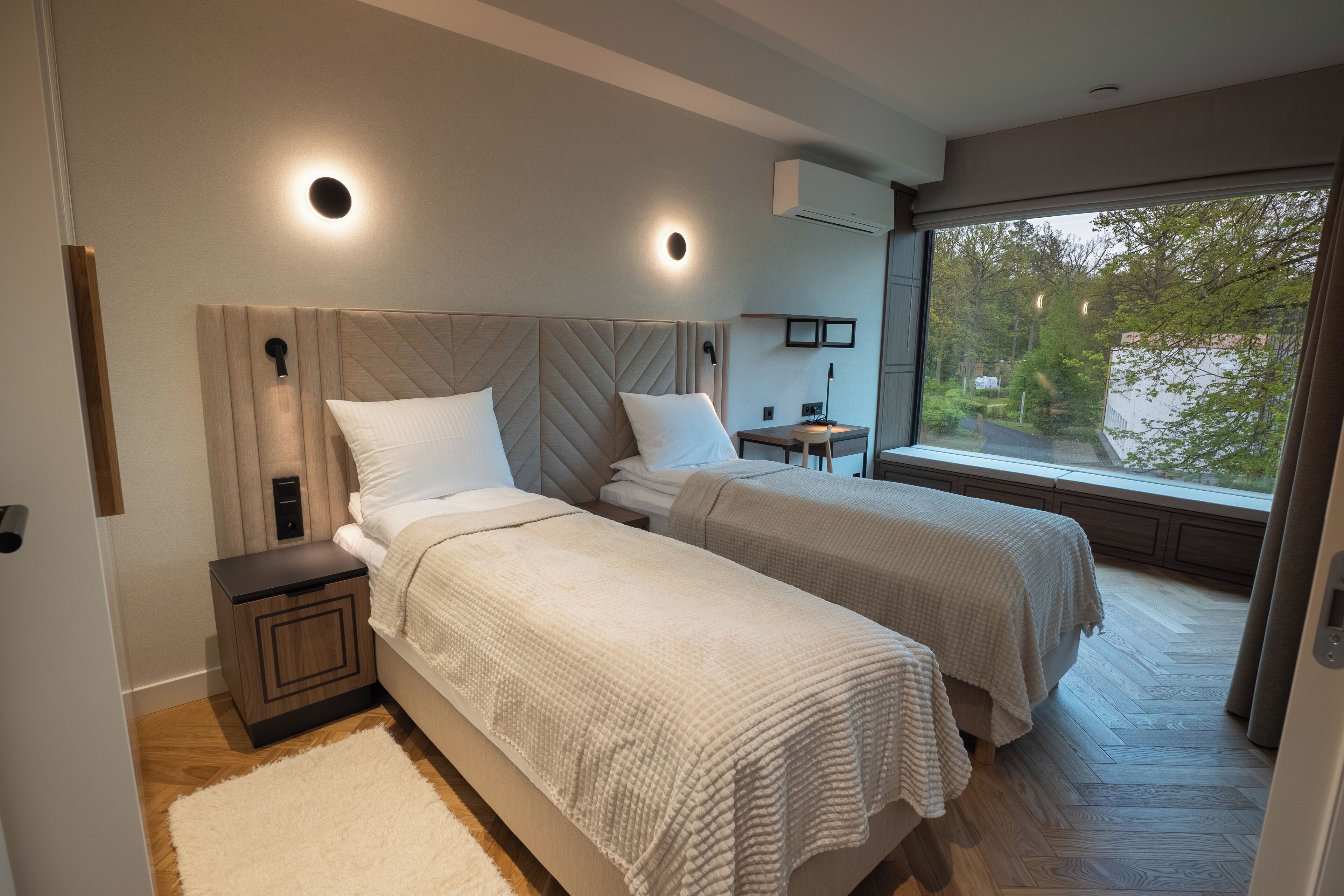 Quarto moderno com duas camas, uma mesa de cabeceira, uma pequena mesa e uma janela grande com vista para área verde.
