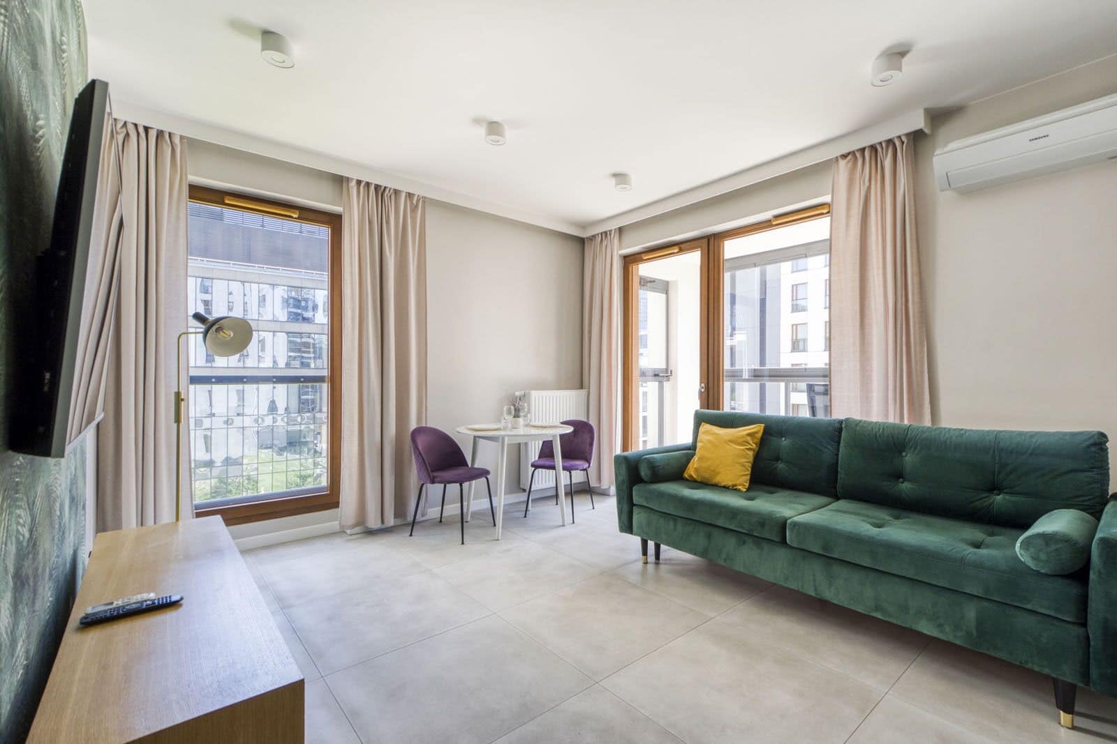Uma sala de estar moderna com um sofá verde, uma mesinha e duas cadeiras. Há janelas amplas com cortinas, um aparelho de ar condicionado na parede e uma TV de tela plana fixada na parede.
