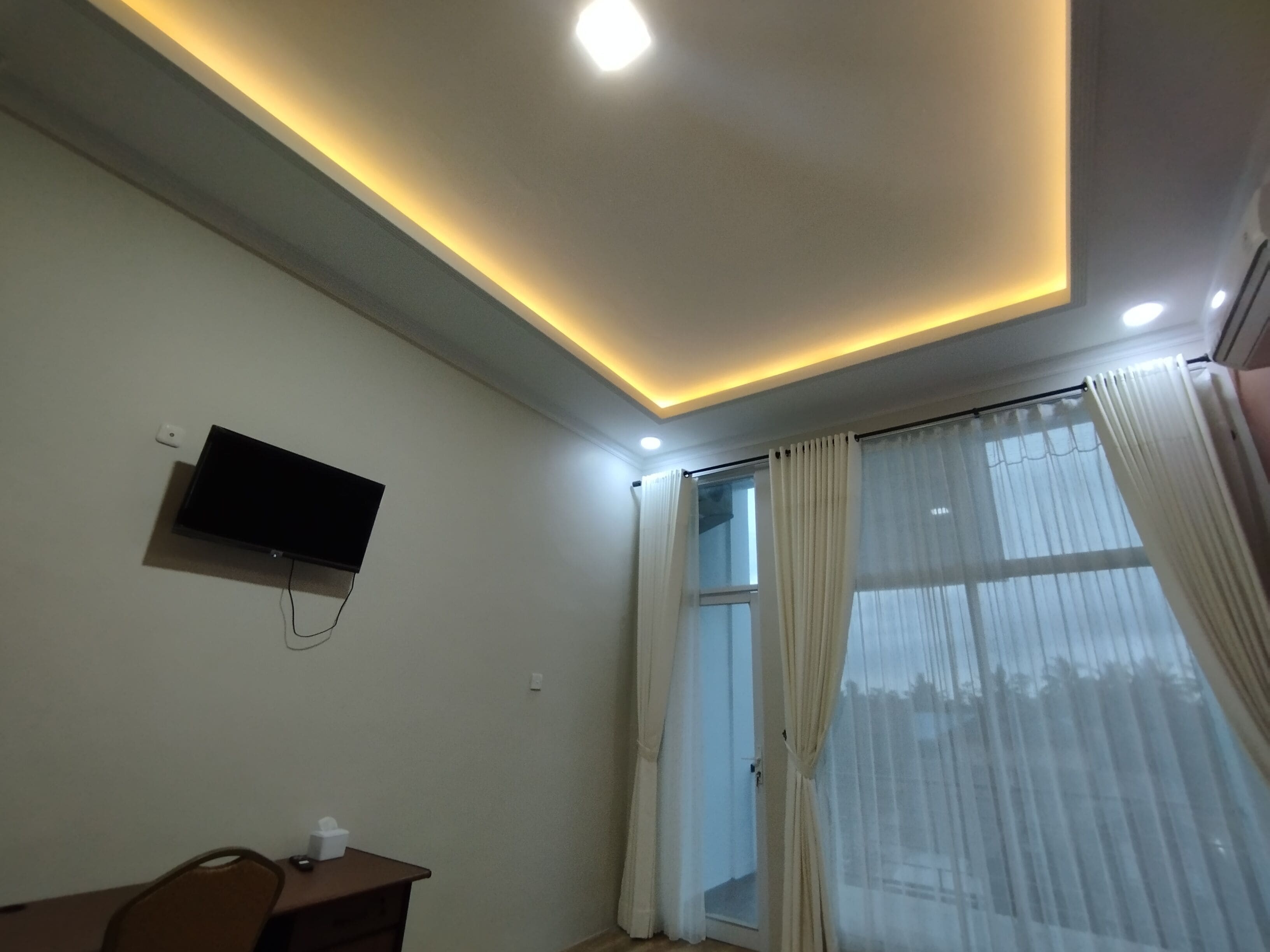 Quarto de hotel com televisão de tela plana na parede, uma escrivaninha com cadeira e uma janela com cortinas finas.