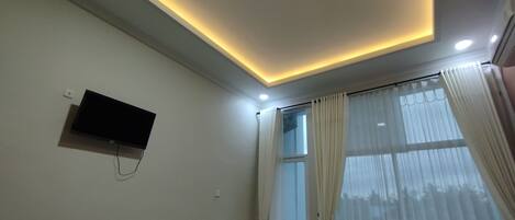 Quarto de hotel com televisão de tela plana na parede, uma escrivaninha com cadeira e uma janela com cortinas finas.