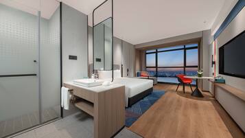 Quarto de hotel moderno com uma cama grande, banheiro com box de vidro, escrivaninha com cadeira e uma janela ampla com vista para a cidade.