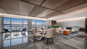 Um hall de hotel moderno com teto alto, grandes janelas com vista para a cidade e uma variedade de áreas de estar, incluindo cadeiras e sofás.