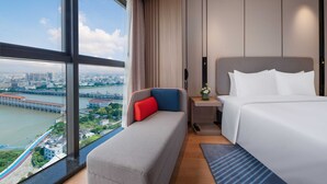 Quarto de hotel moderno com uma grande janela que oferece vista para a cidade, uma cama, um sofá e uma pequena mesa de cabeceira com uma luminária.