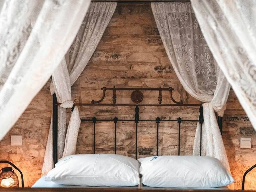 Uma cama com almofadas brancas e cabeceira de metal, apoiada em uma parede de pedra com teto de madeira.