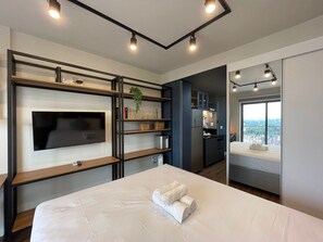 Quarto de hotel moderno com cama, televisão de tela plana, estantes embutidas, cozinha e varanda com vista para a cidade.