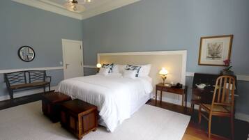 Quarto com cama grande, mesa de cabeceira com luminária, cadeira, banqueta e quadro na parede.