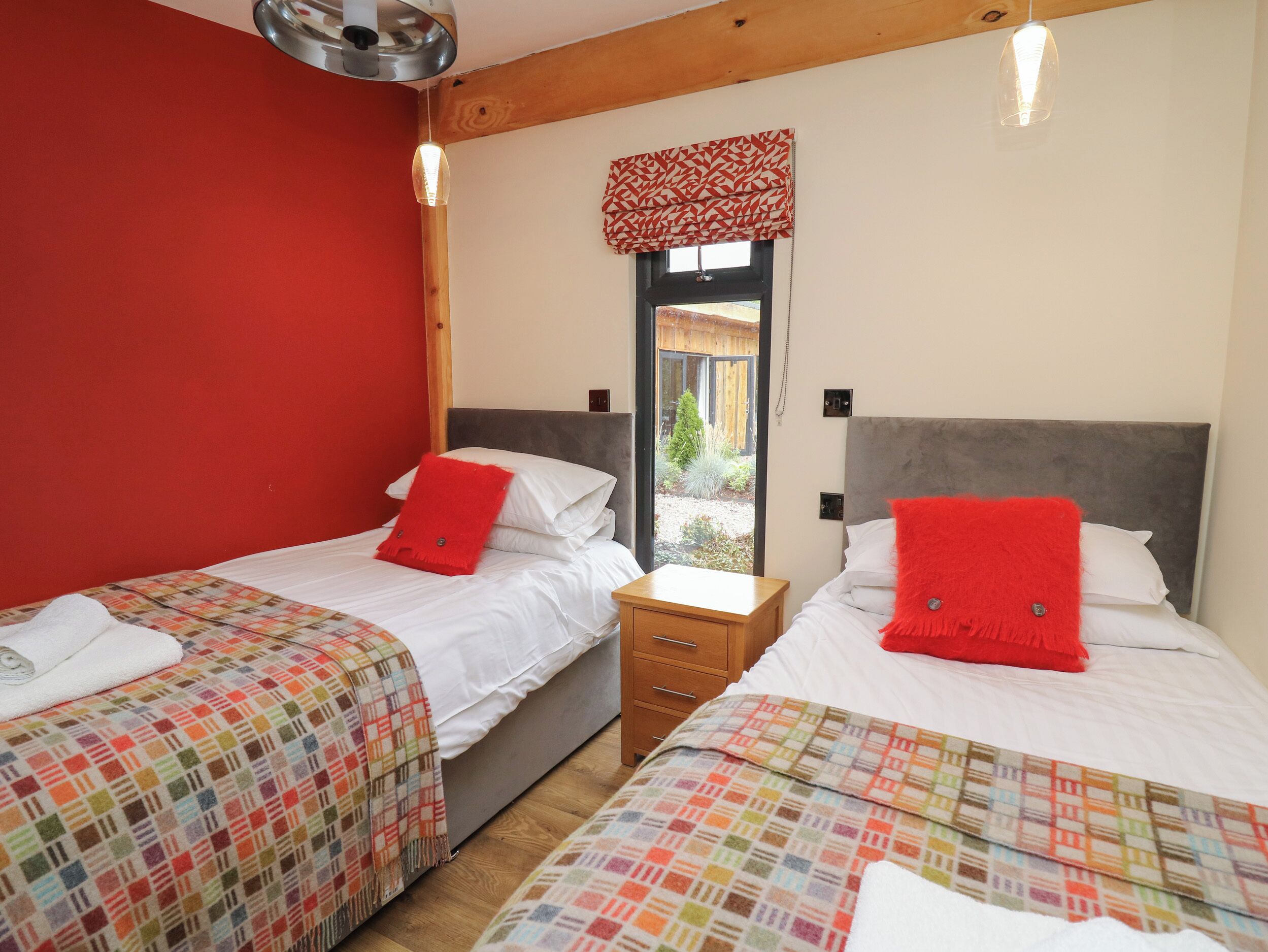 Quarto com duas camas, cada uma com travesseiro vermelho e manta xadrez colorida. Há uma mesinha de cabeceira de madeira entre as camas e uma janela com cortina vermelha estampada.