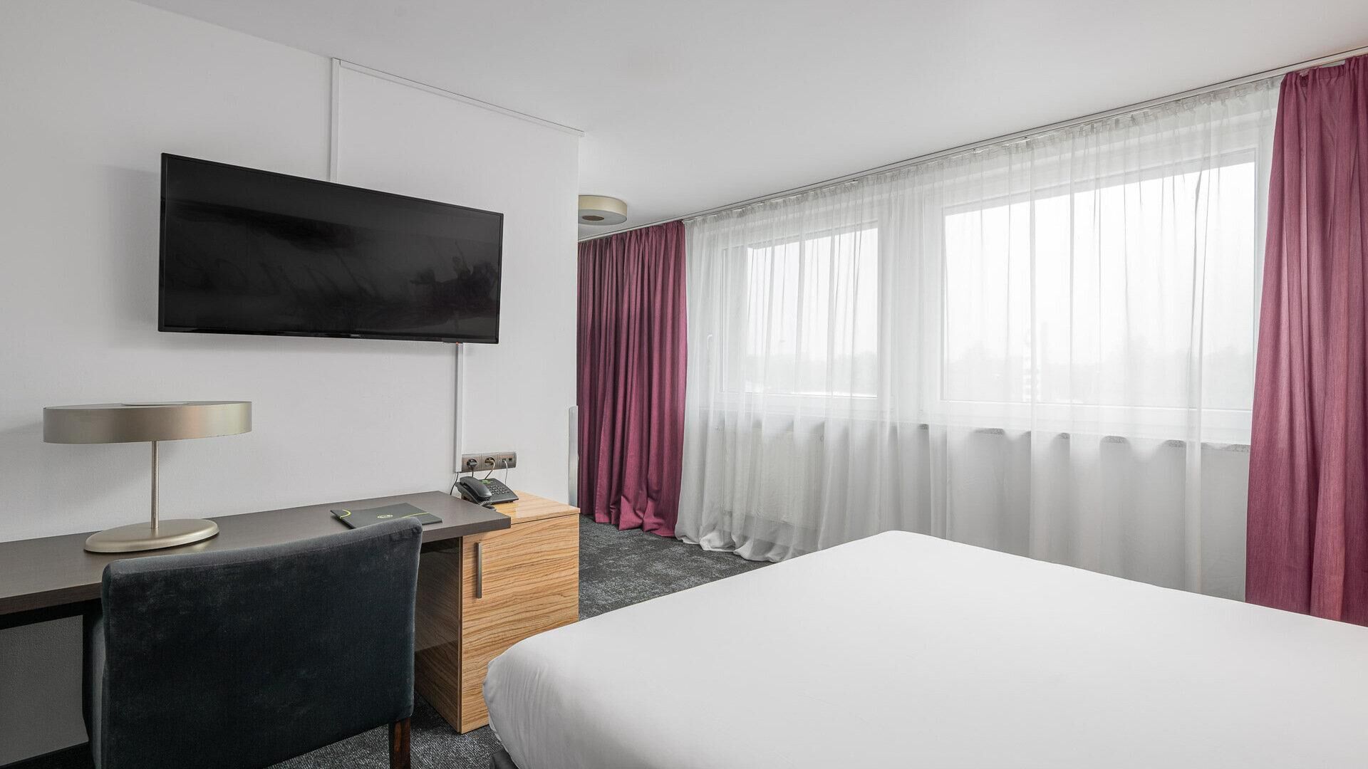 Quarto de hotel com cama, escrivaninha com cadeira, TV de tela plana, abajur e janela com cortinas brancas translúcidas e um tecido vermelho.