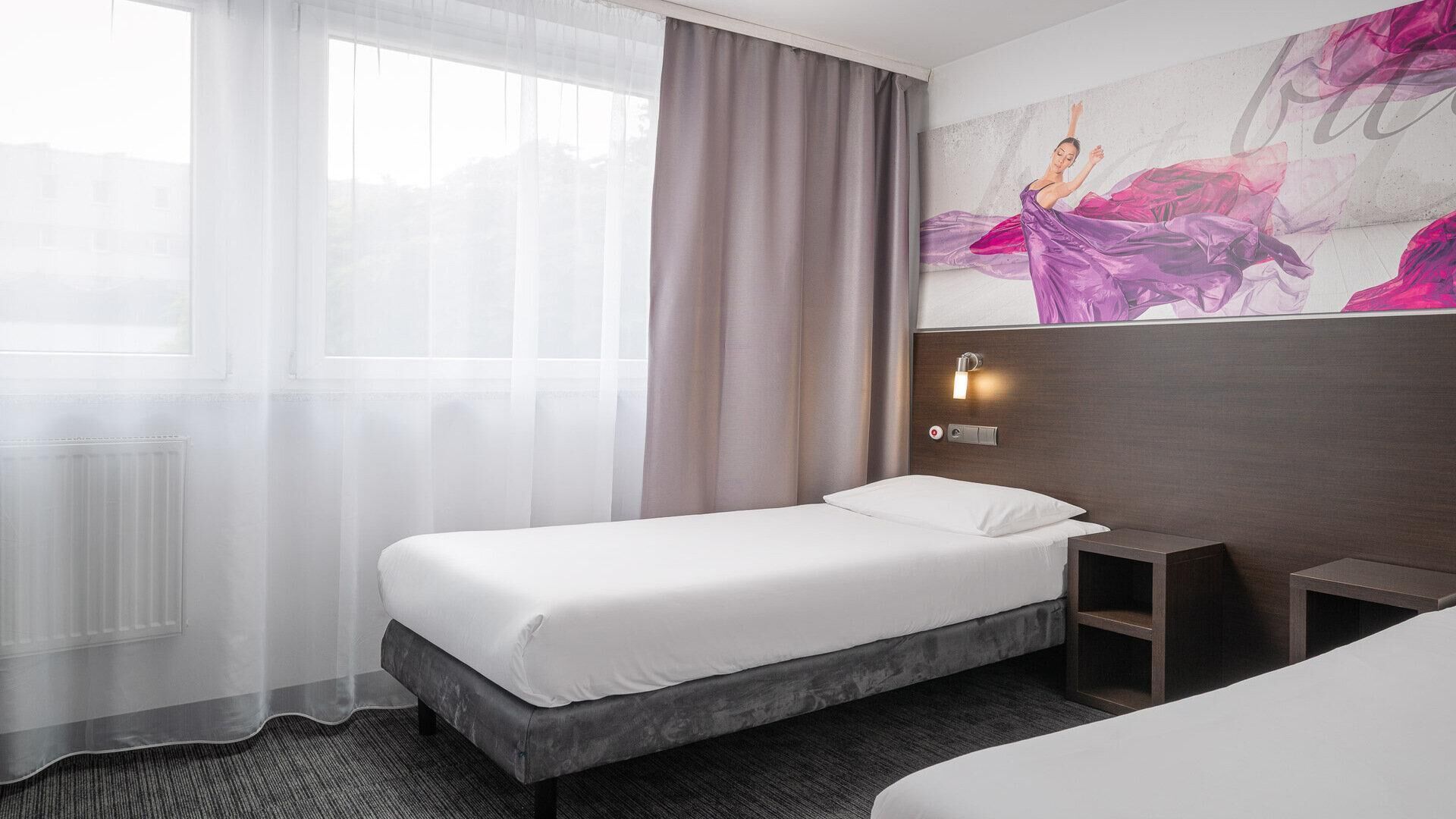 Quarto de hotel com cama, mesa de cabeceira, luminária na parede e janela com cortinas translúcidas.