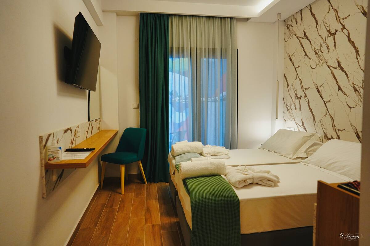 Quarto de hotel com cama, escrivaninha com cadeira, televisão fixada na parede e janela com cortinas.