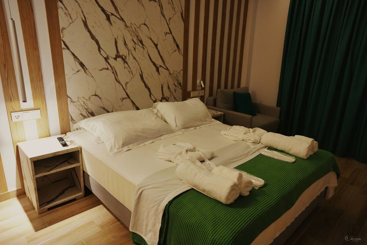 Uma cama bem arrumada com lençóis brancos e um cobertor verde, um criado-mudo de madeira com um controle remoto e uma cortina verde.