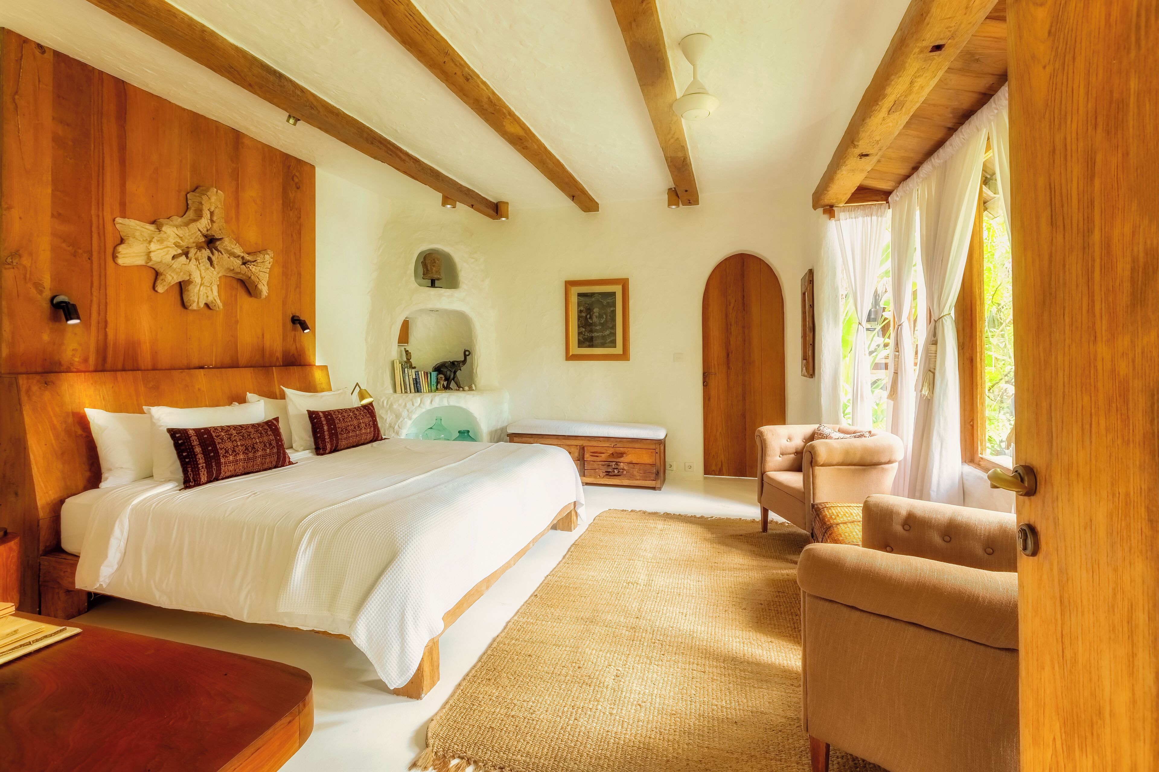 Quarto com uma cama grande, vigas de madeira, cabeceira de madeira, mesa de cabeceira, uma cadeira e uma janela com cortinas.