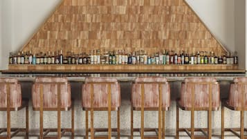 Uma área de bar com teto de madeira inclinado e uma longa estante com diversas garrafas de bebidas. Há seis banquetas com assentos de fibra.