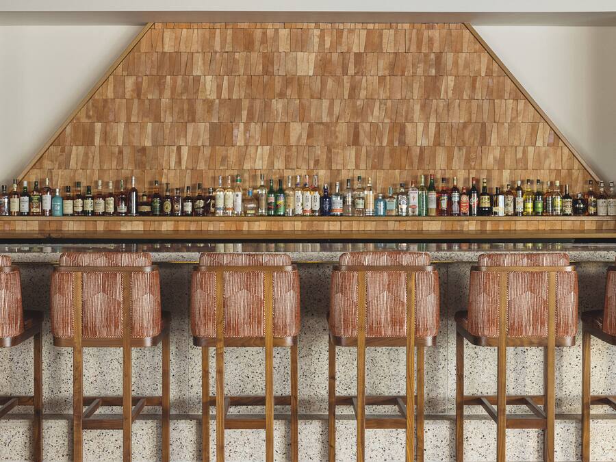Uma área de bar com teto de madeira inclinado e uma longa estante com diversas garrafas de bebidas. Há seis banquetas com assentos de fibra.