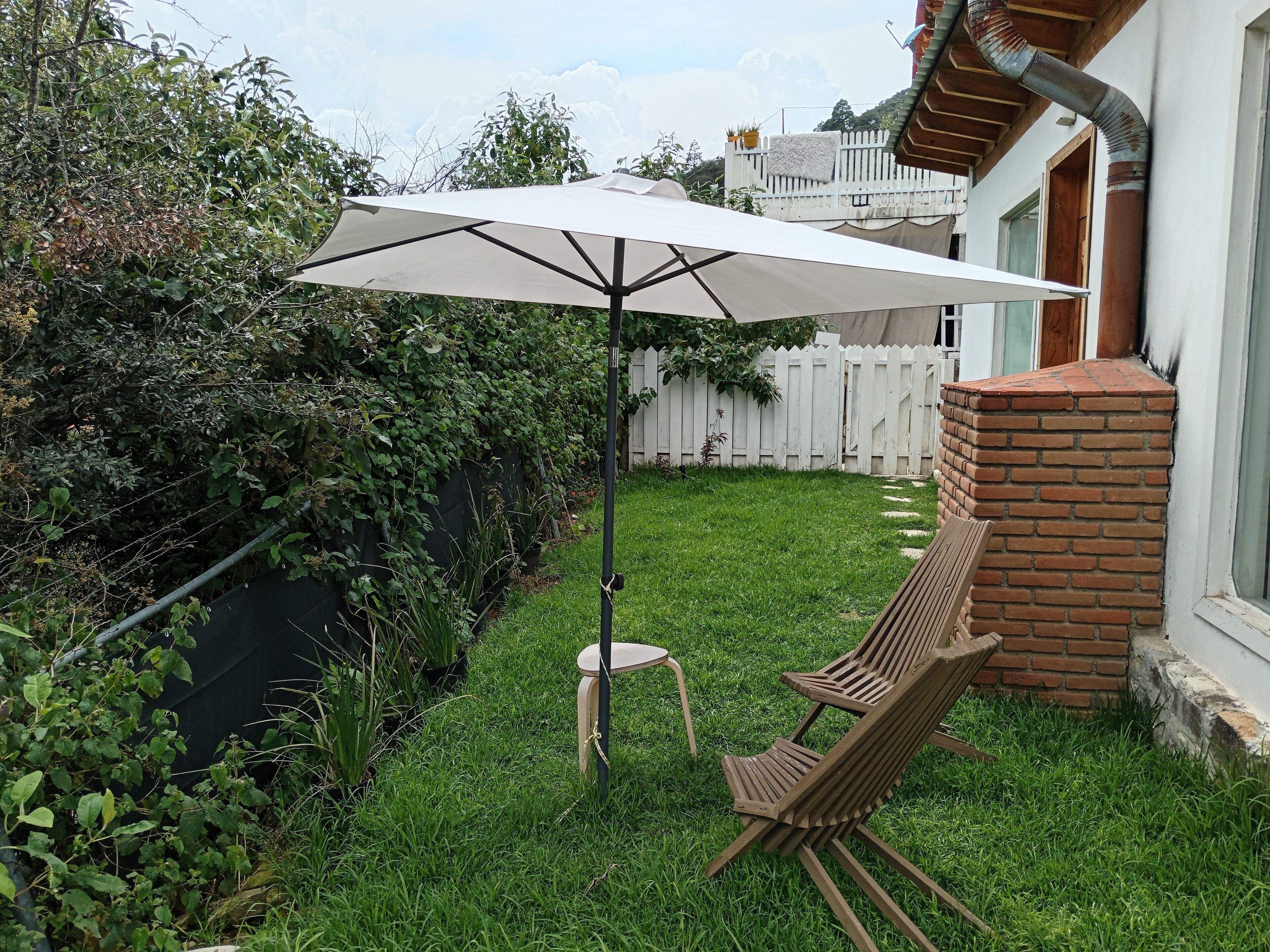 Um quintal com um guarda-sol branco, uma chaminé de tijolos e uma cadeira de madeira.