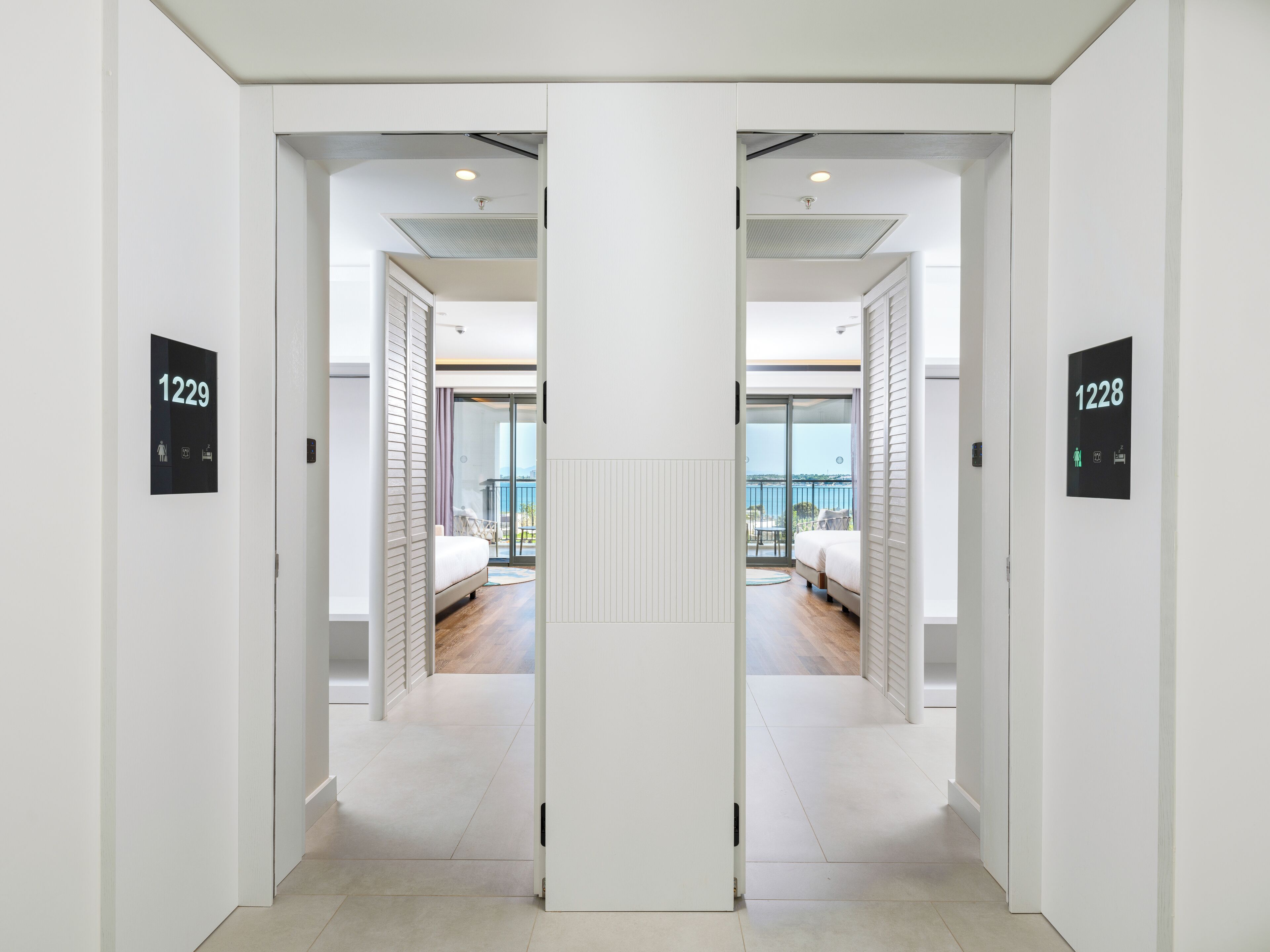 Corredor de hotel com portas identificadas como 1229 e 1228, que dão acesso a quartos com camas, vista para o mar e varanda.