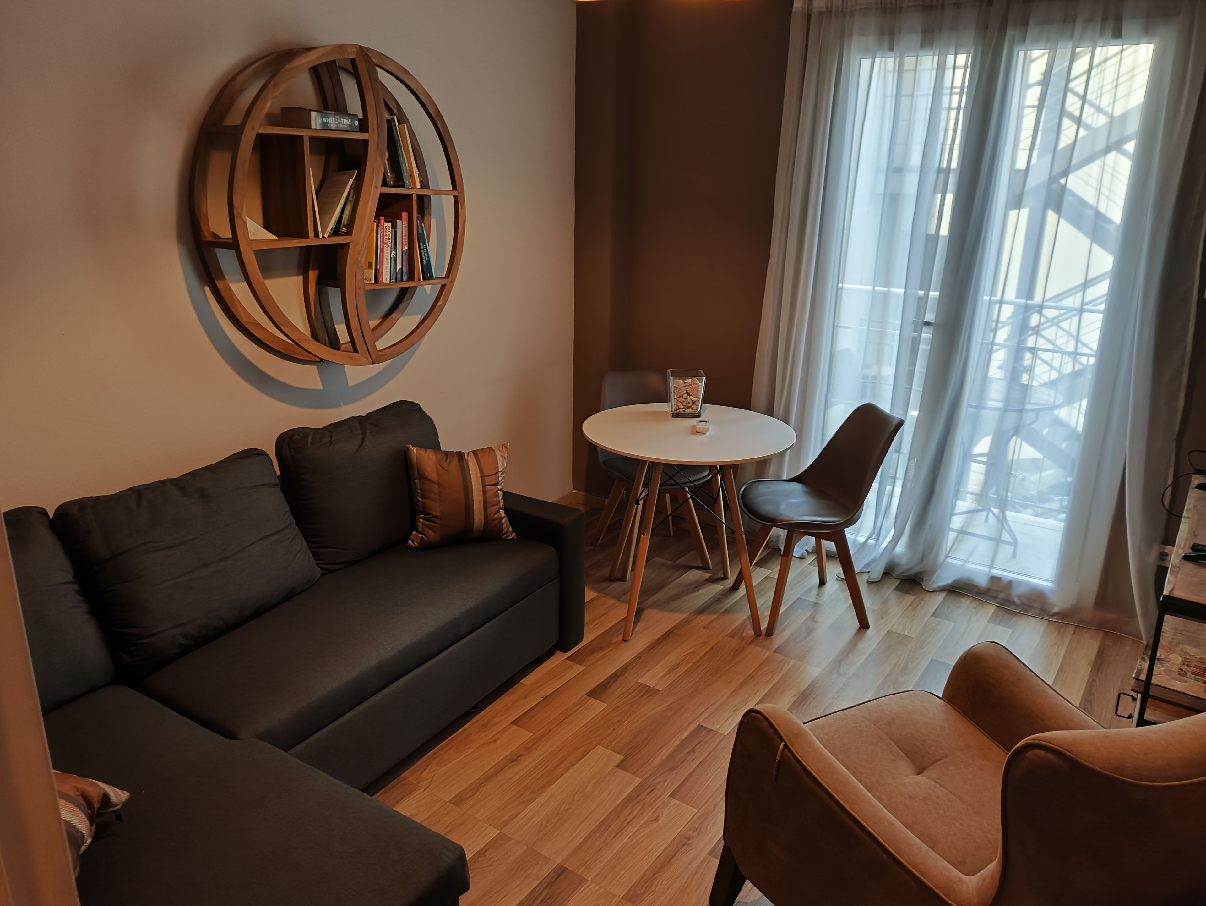 Uma sala de estar moderna com sofá, estante de madeira redonda, mesa redonda com cadeiras e uma grande janela com cortinas.