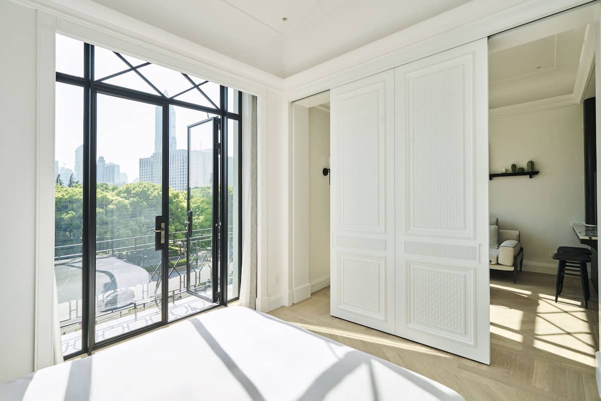 Quarto de hotel moderno com uma grande porta de vidro deslizante para uma varanda, um guarda-roupa branco e vista para a paisagem urbana.