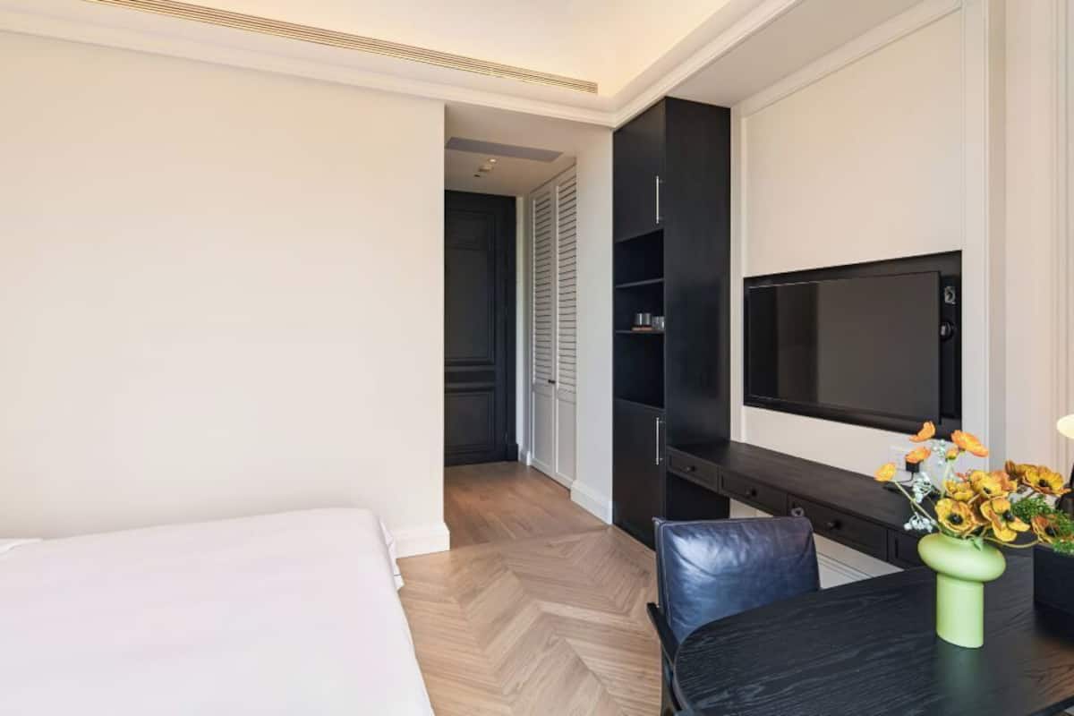 Quarto de hotel com uma cama, televisão, escrivaninha com um vaso de flores e vista para o corredor.
