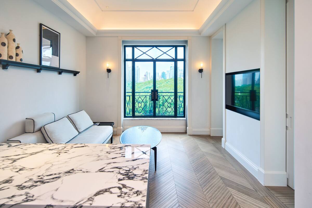Uma sala de estar moderna com uma mesa de centro de mármore, um sofá, uma TV na parede e uma janela grande com vista para a cidade.