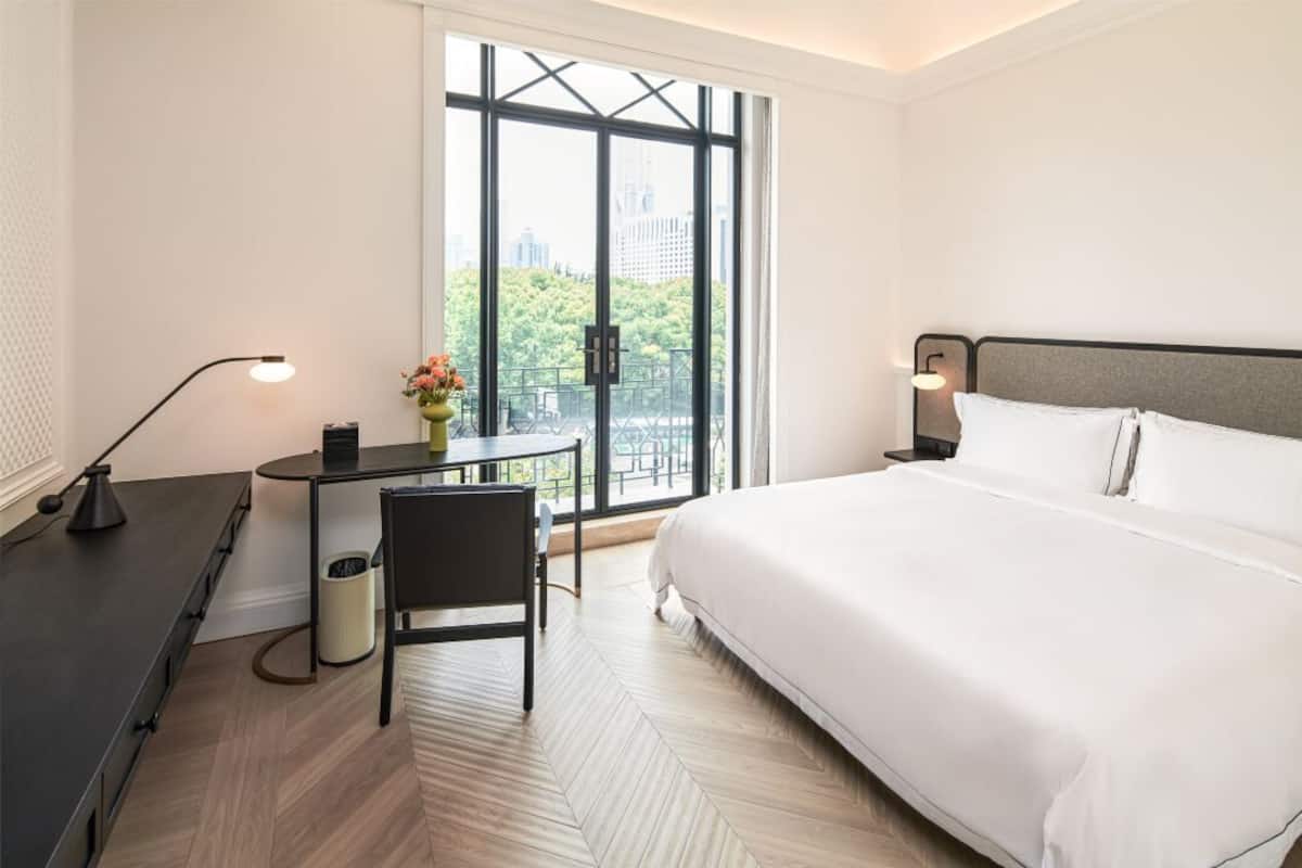 Quarto de hotel com uma cama grande, uma escrivaninha com cadeira, uma mesinha com um vaso e vista para a cidade através de janelas amplas.