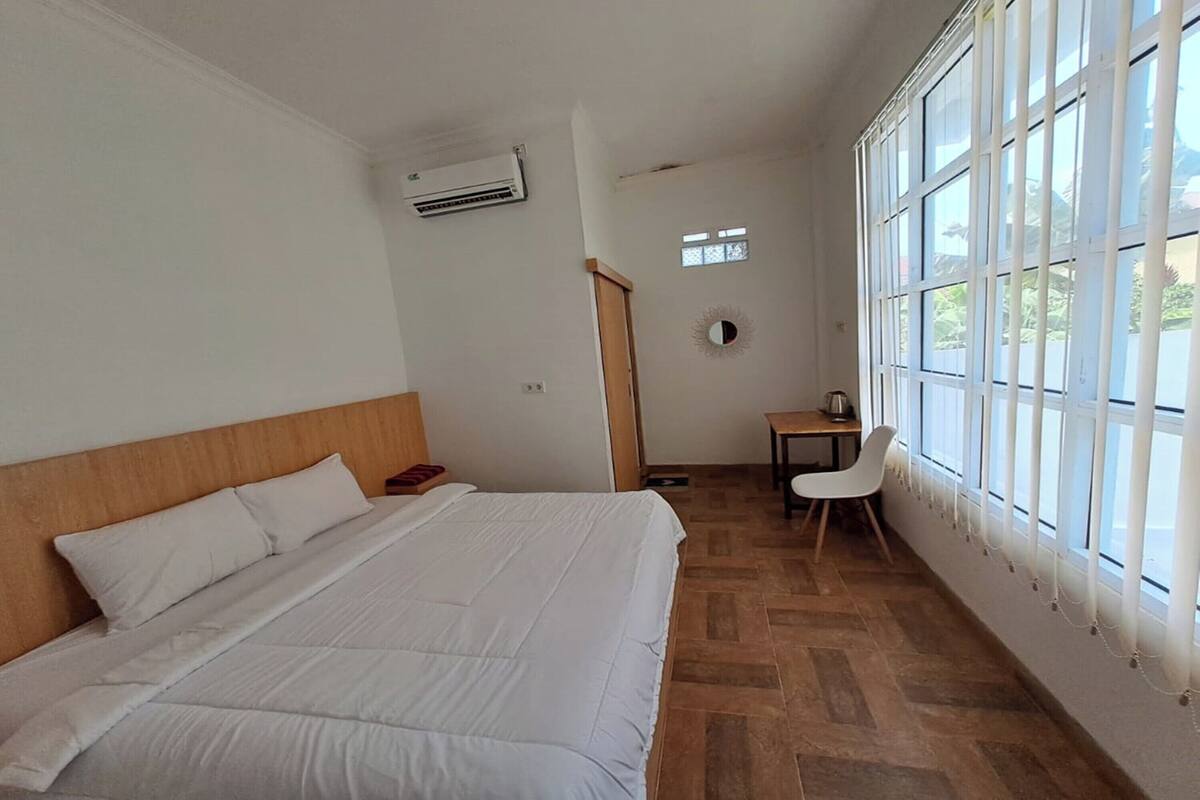 Quarto de hotel com uma cama grande, uma mesa e uma cadeira. Há uma janela com persianas e um aparelho de ar condicionado na parede.
