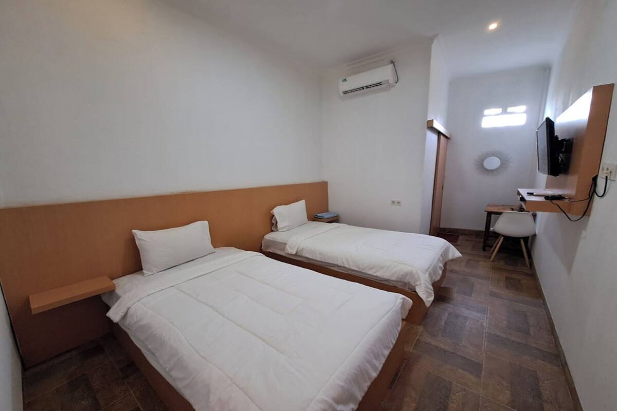 Quarto de hotel com duas camas de solteiro, uma escrivaninha, uma cadeira, um aparelho de ar condicionado e uma televisão fixada na parede.