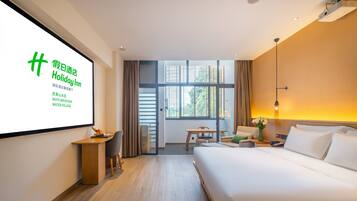 Quarto de hotel com cama, escrivaninha, cadeira, mesinha e uma janela grande.