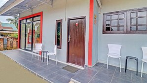 Um edifício com entrada emoldurada em vermelho, uma pequena área coberta com cadeiras e uma janela com padrão de grade.