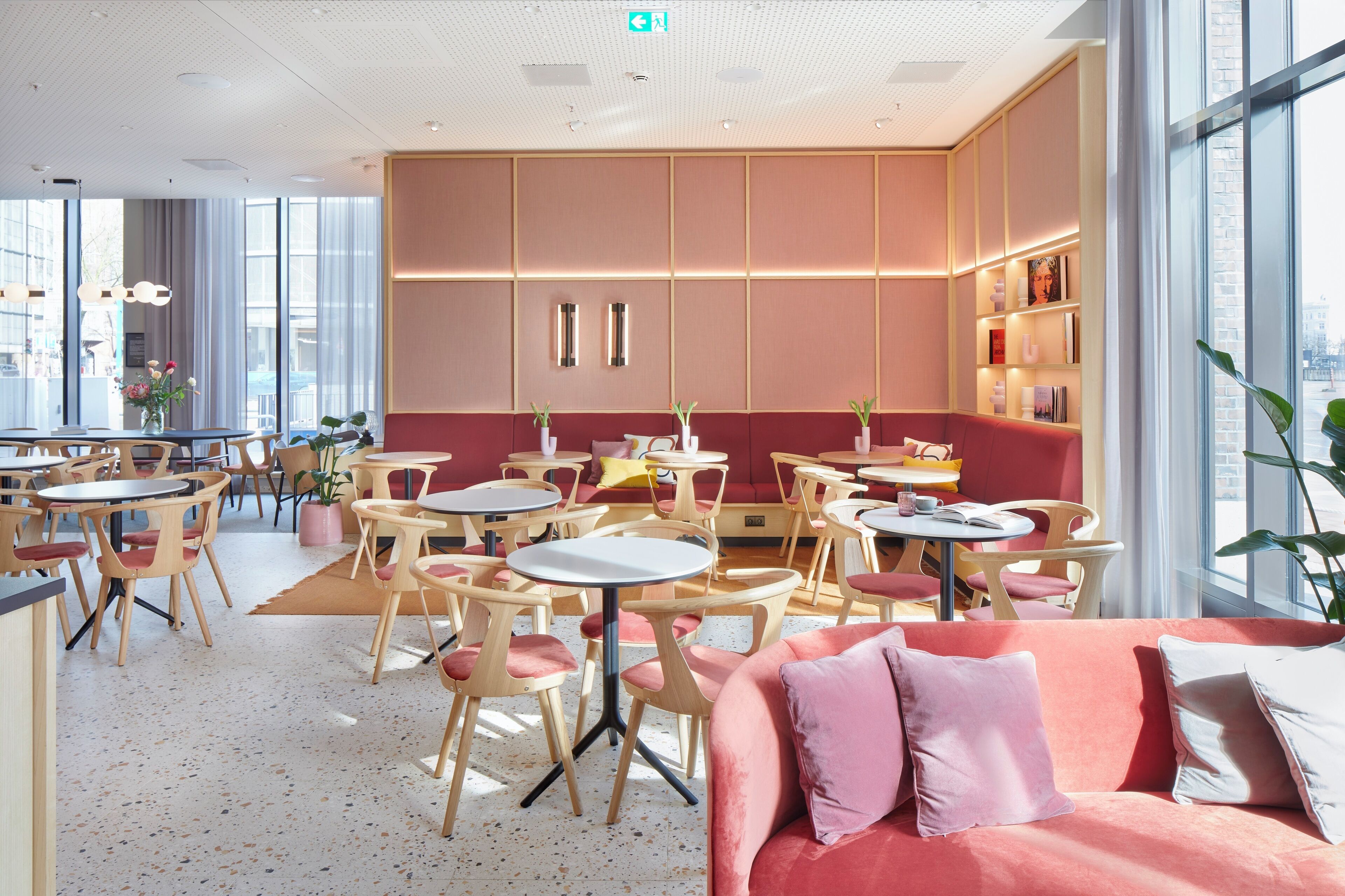 Um café moderno com mesas e cadeiras redondas, um sofá vermelho e amplas janelas com vista para a cidade.