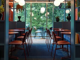 Restaurante com várias mesas e cadeiras, grandes janelas com vista para árvores e luminárias pendentes esféricas.