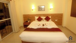 Quarto de hotel com uma cama grande, cabeceira de madeira, duas luzes fixadas na parede, uma mesinha com uma planta e uma porta de vidro deslizante para uma varanda.