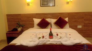 Quarto de hotel com cabeceira de madeira, duas almofadas vermelhas, uma garrafa de champanhe e um buquê de rosas na cama.