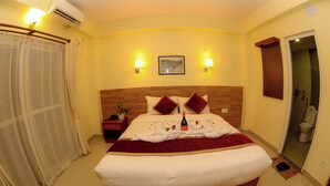 Quarto de hotel com uma cama grande, cabeceira de madeira, duas luminárias fixadas na parede, um quadro emoldurado e um banheiro visível através de uma porta aberta.