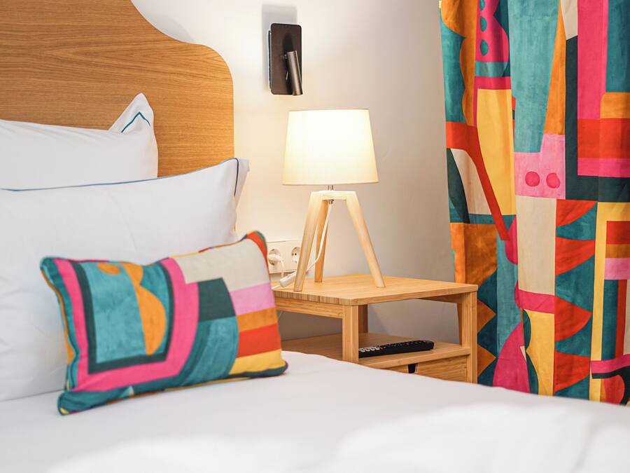 Cama bem arrumada com lençóis brancos, um travesseiro colorido, mesa de cabeceira de madeira com abajur e cortina com padrão geométrico.