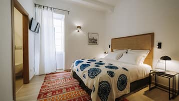 Quarto com cama, mesa de cabeceira, televisão e janela com cortinas.