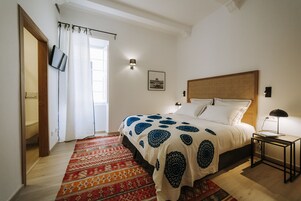 Quarto com cama, mesa de cabeceira, televisão e janela com cortinas.