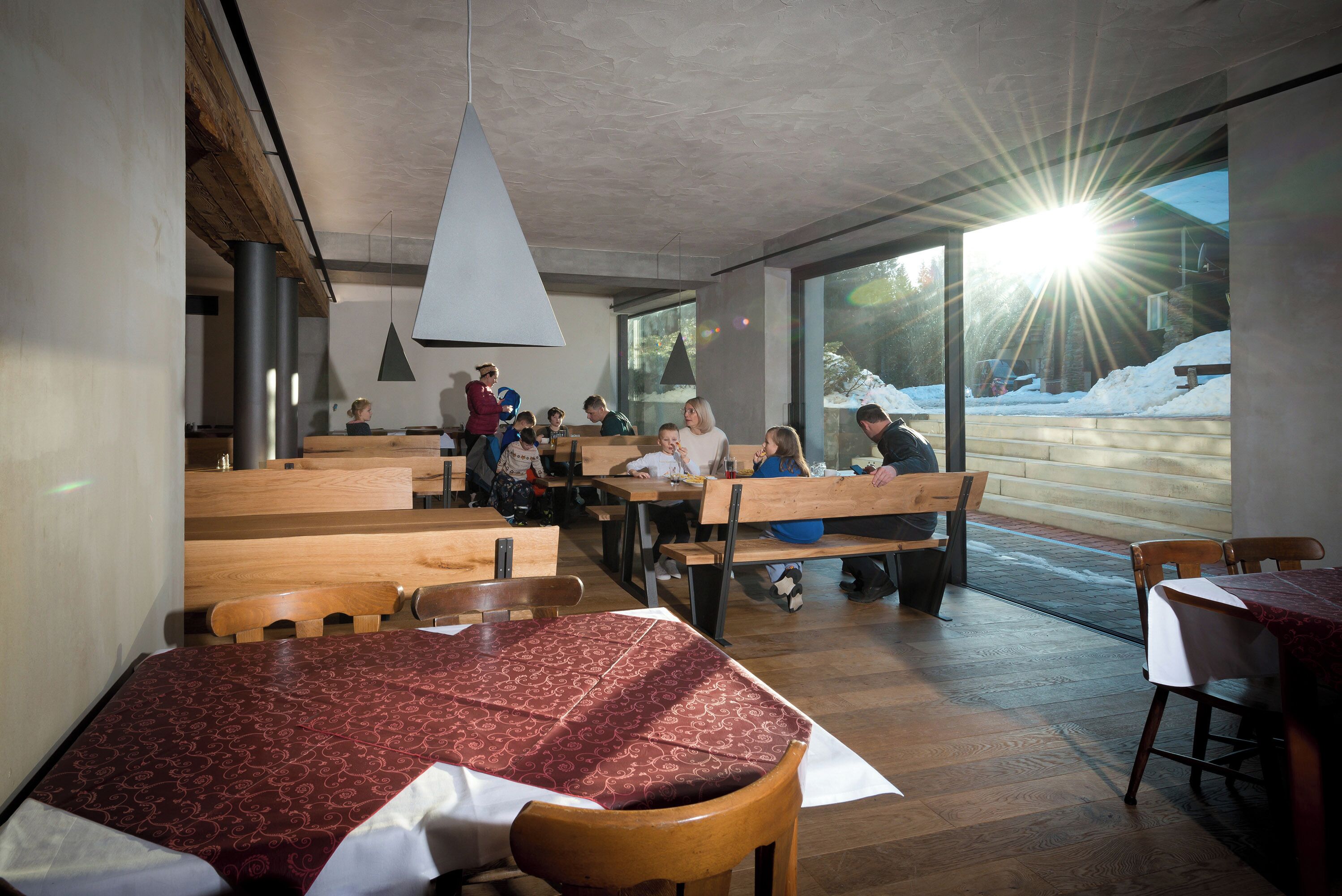Um restaurante com mesas e bancos de madeira, janelas amplas e vista para uma área externa coberta de neve.