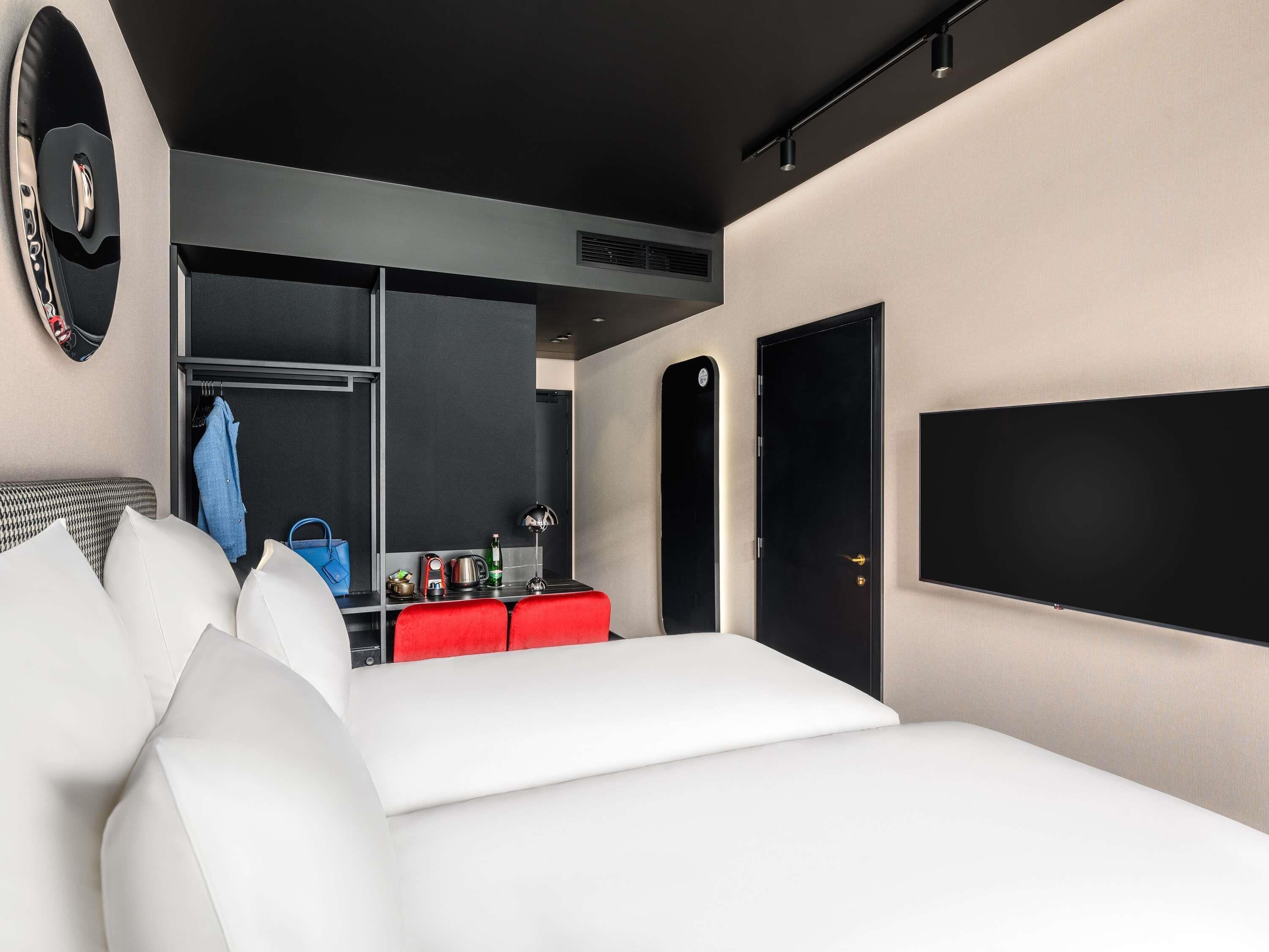 Quarto de hotel moderno com uma cama grande, uma escrivaninha com cadeira, um guarda-roupa, um espelho e uma televisão fixada na parede.