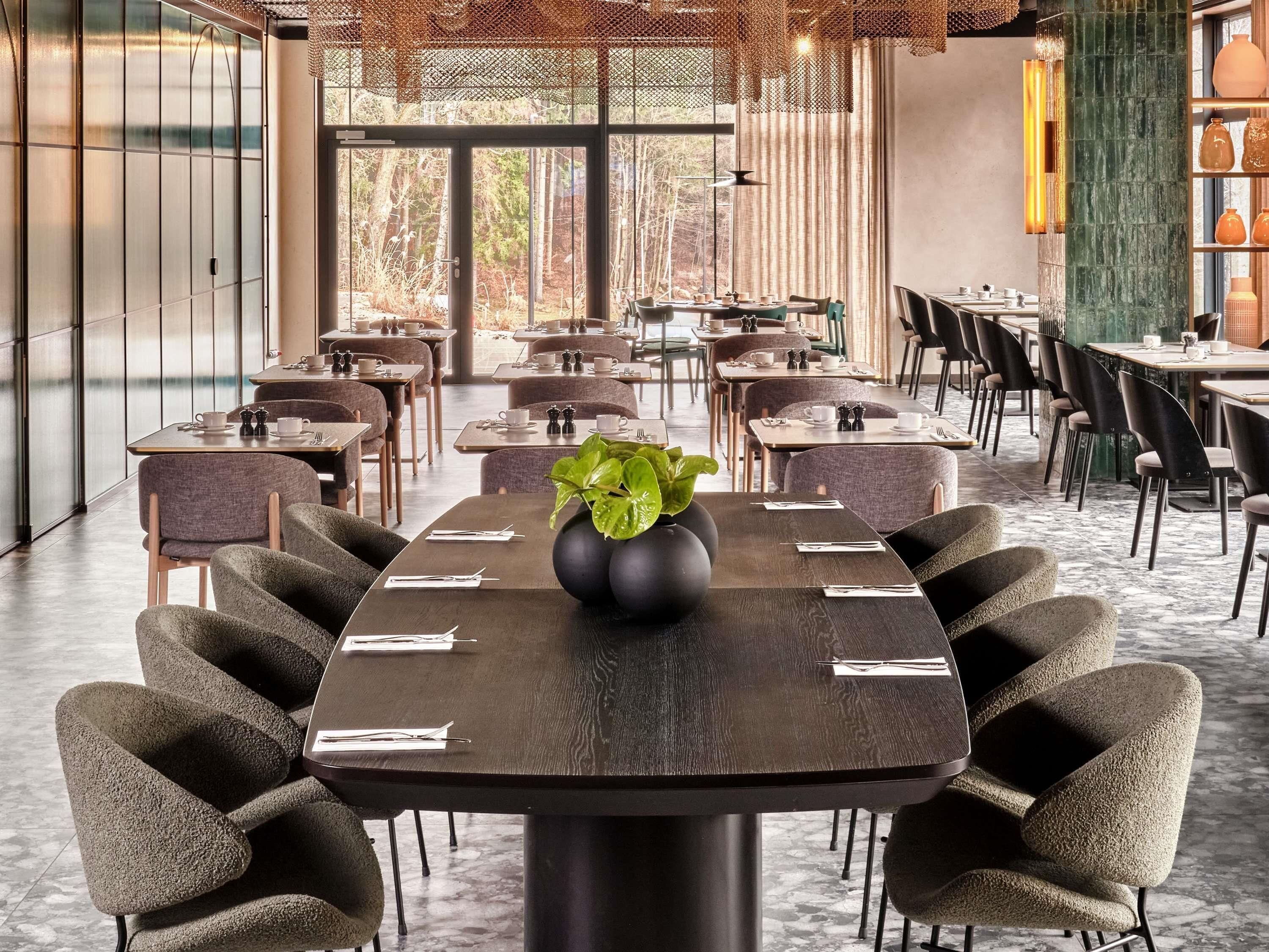 Uma área de refeições moderna com mesas redondas, cadeiras confortáveis e uma mesa central com uma planta verde.