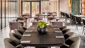 Uma área de refeições moderna com mesas redondas, cadeiras confortáveis e uma mesa central com uma planta verde.