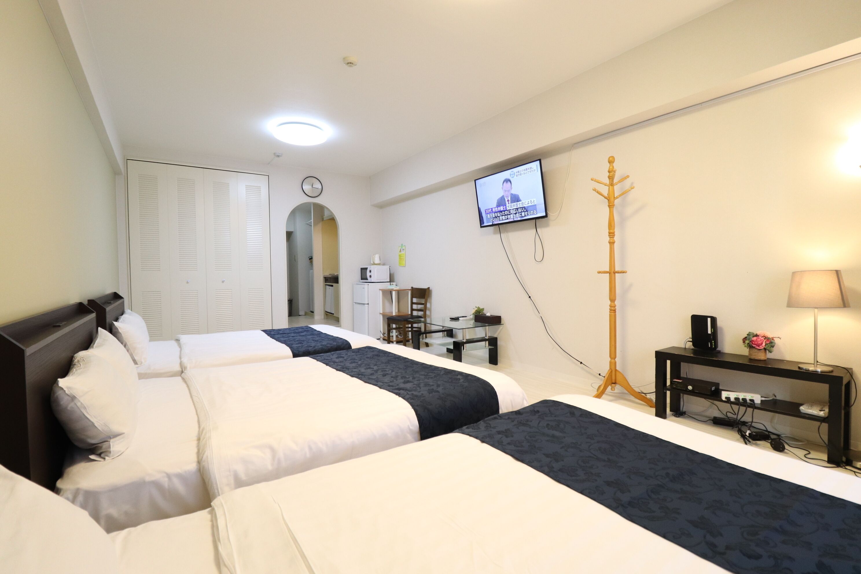 Quarto de hotel com três camas, uma escrivaninha, uma televisão e um abajur.