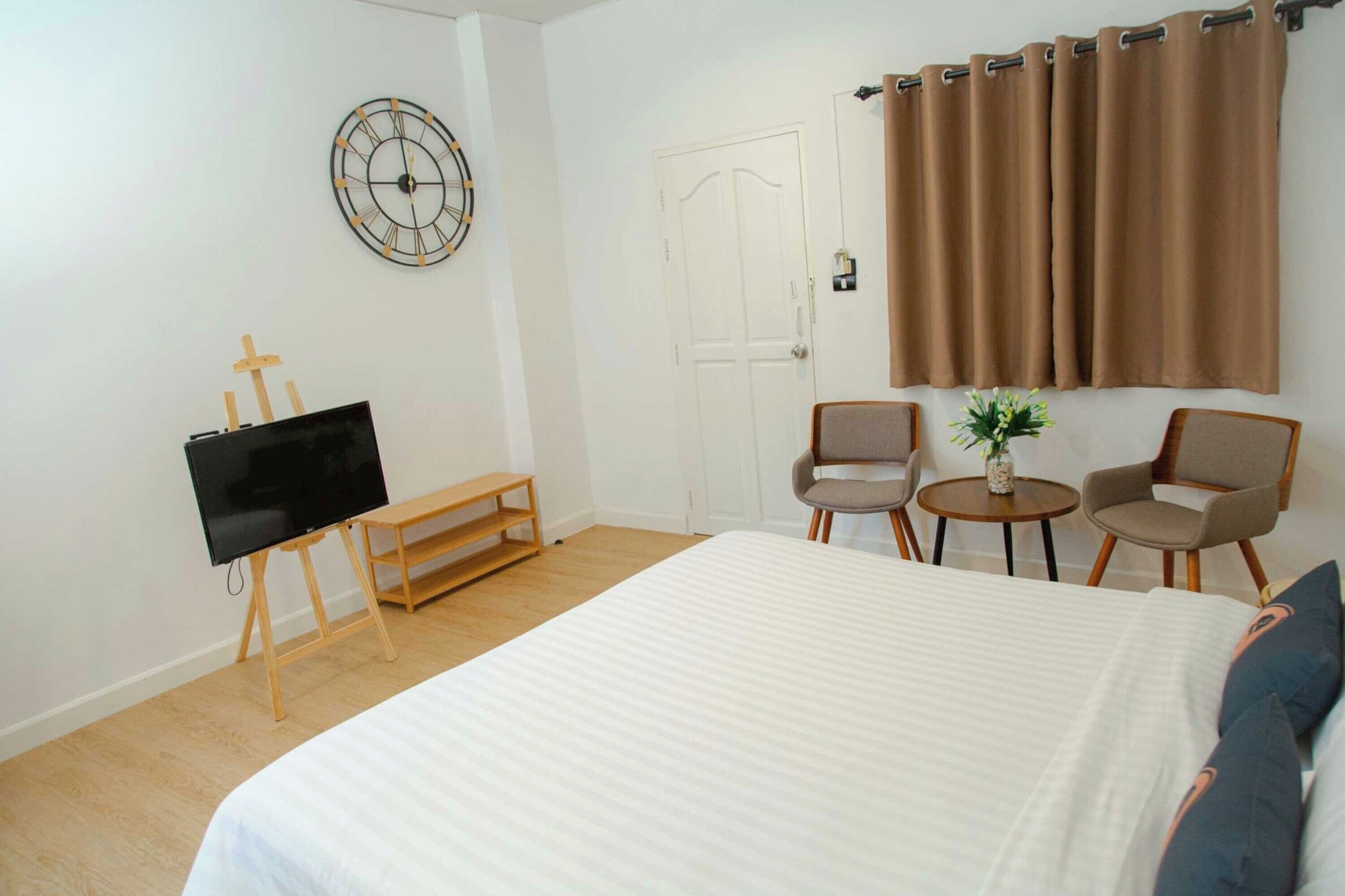 Quarto com cama, televisão em móvel, duas cadeiras, pequena mesa com planta e porta.