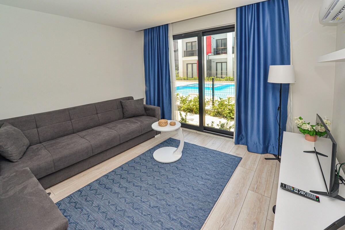 Uma sala de estar moderna com um sofá cinza, uma mesinha de apoio branca, um tapete azul estampado e vista para uma piscina através de uma porta de vidro.