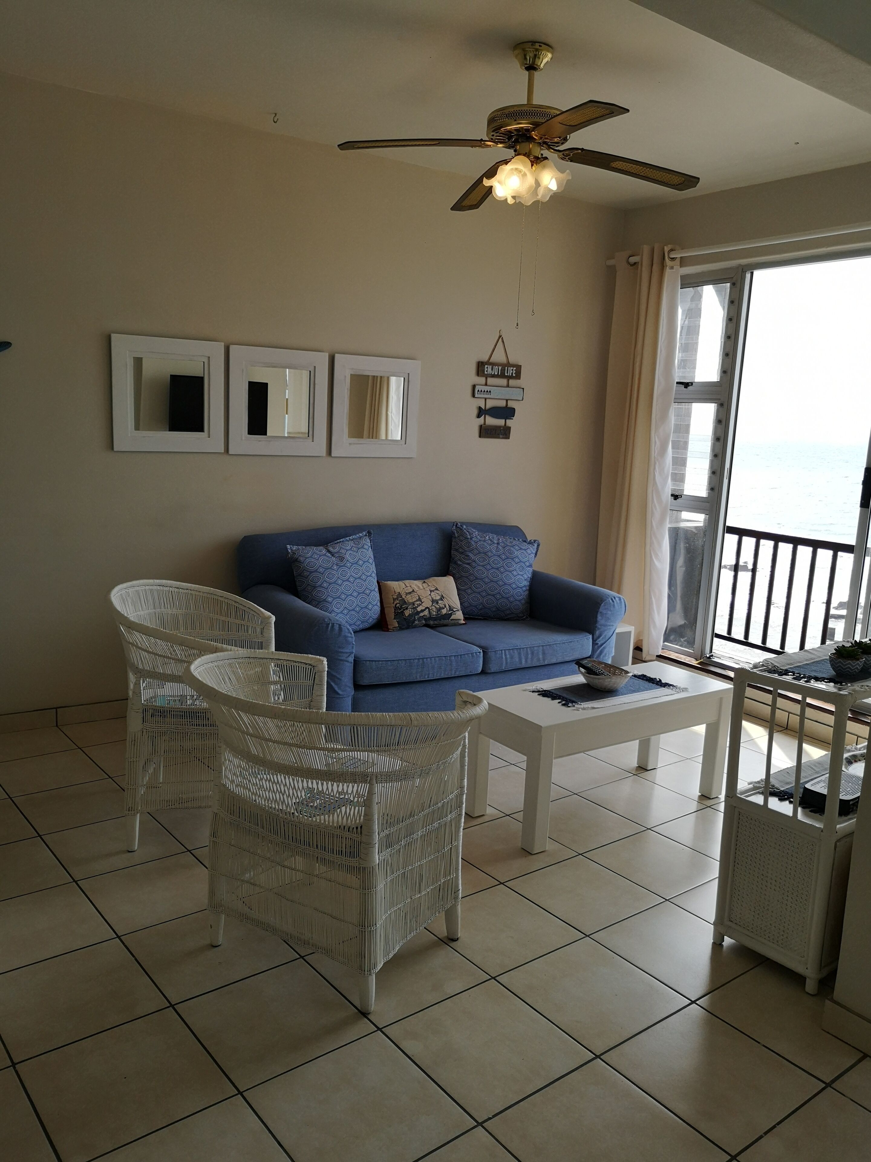 Uma sala de estar com um sofá azul, cadeiras de vime branco e uma mesa de centro branca, além de um ventilador de teto.