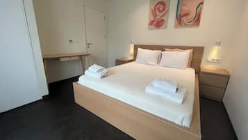 Cama bem arrumada com lençóis e toalhas brancas, cabeceira de madeira, duas mesas de cabeceira com abajures e dois quadros abstratos na parede.
