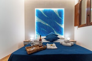 Cama bem arrumada com colcha azul, uma garrafa de vinho, copos e toalhas sobre uma mesa de cabeceira.